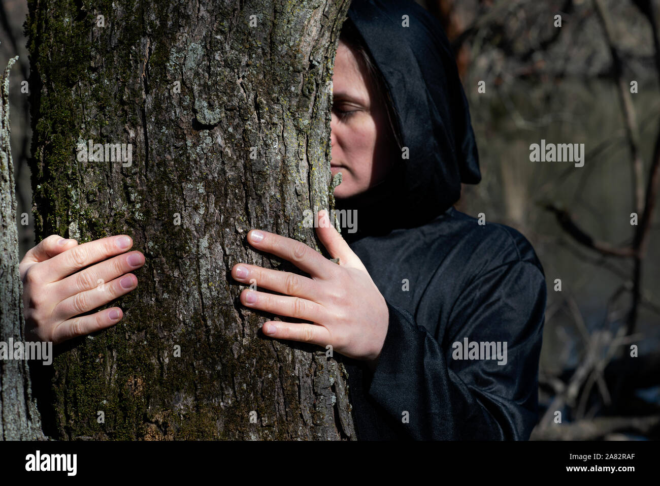 Close-up of Hooded Woman in black robes embracing a Tree Trunk in the ...
