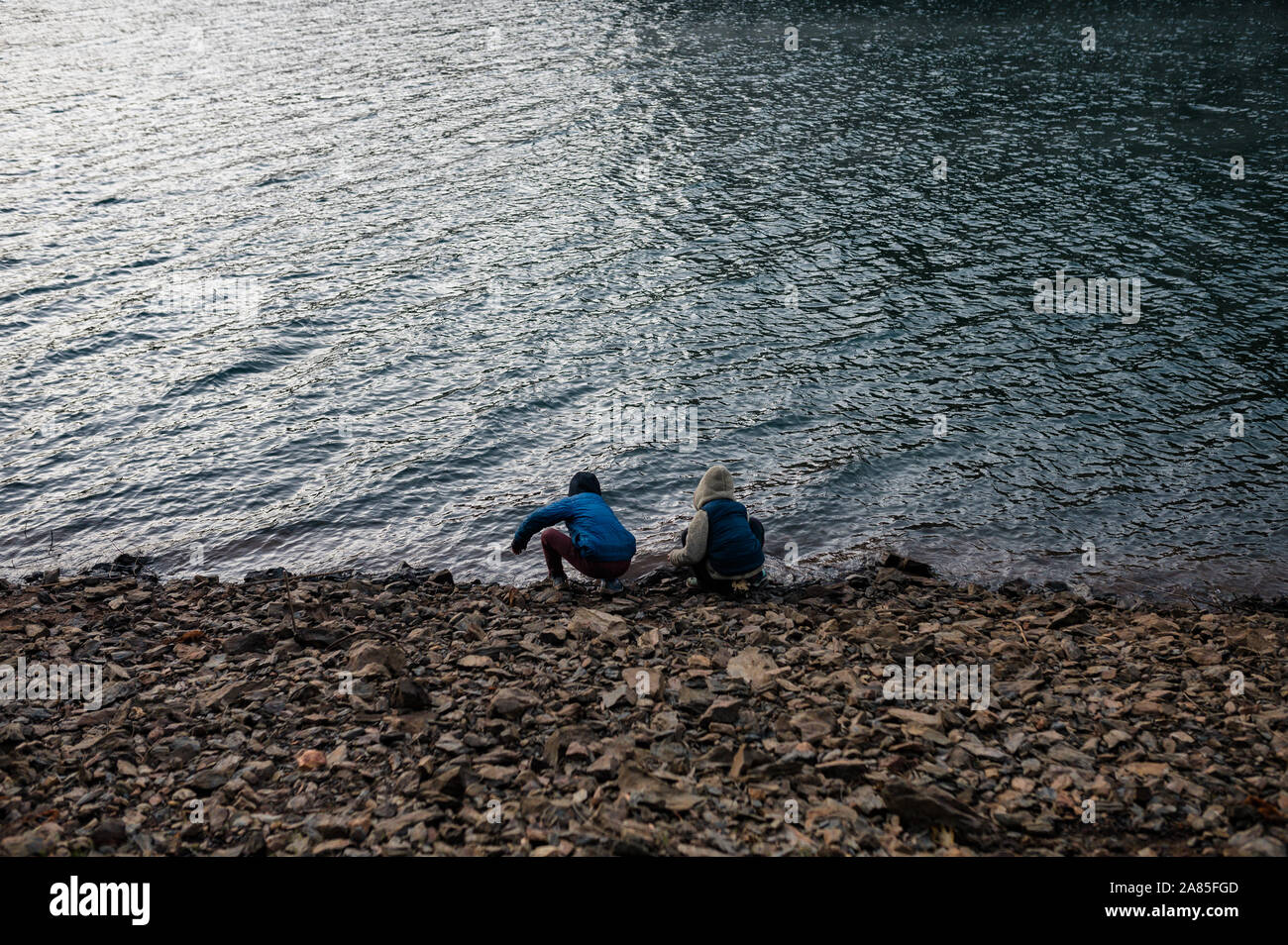 Two children squatting on rocks near the edge of a lake Stock Photo - Alamy