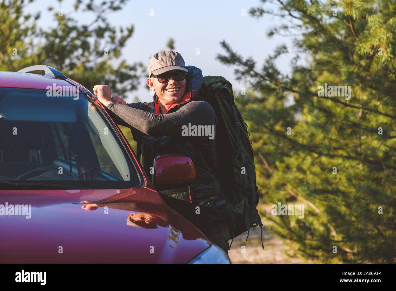 Happy senior tourist with a backpack pose near red car in forest. A ...