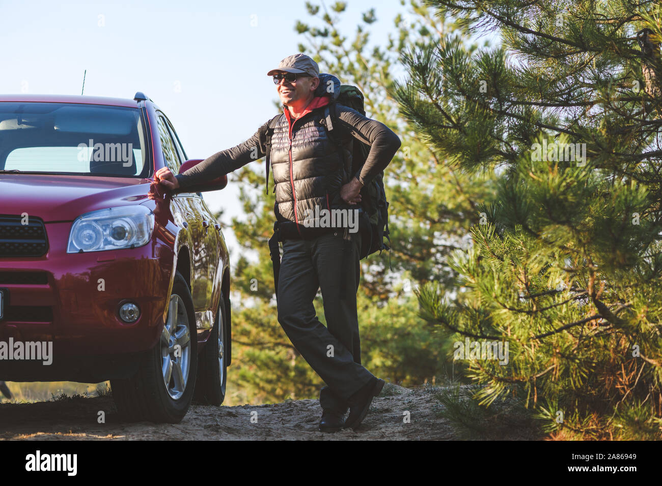 Happy senior tourist with a backpack lean on red car in forest. Elderly ...