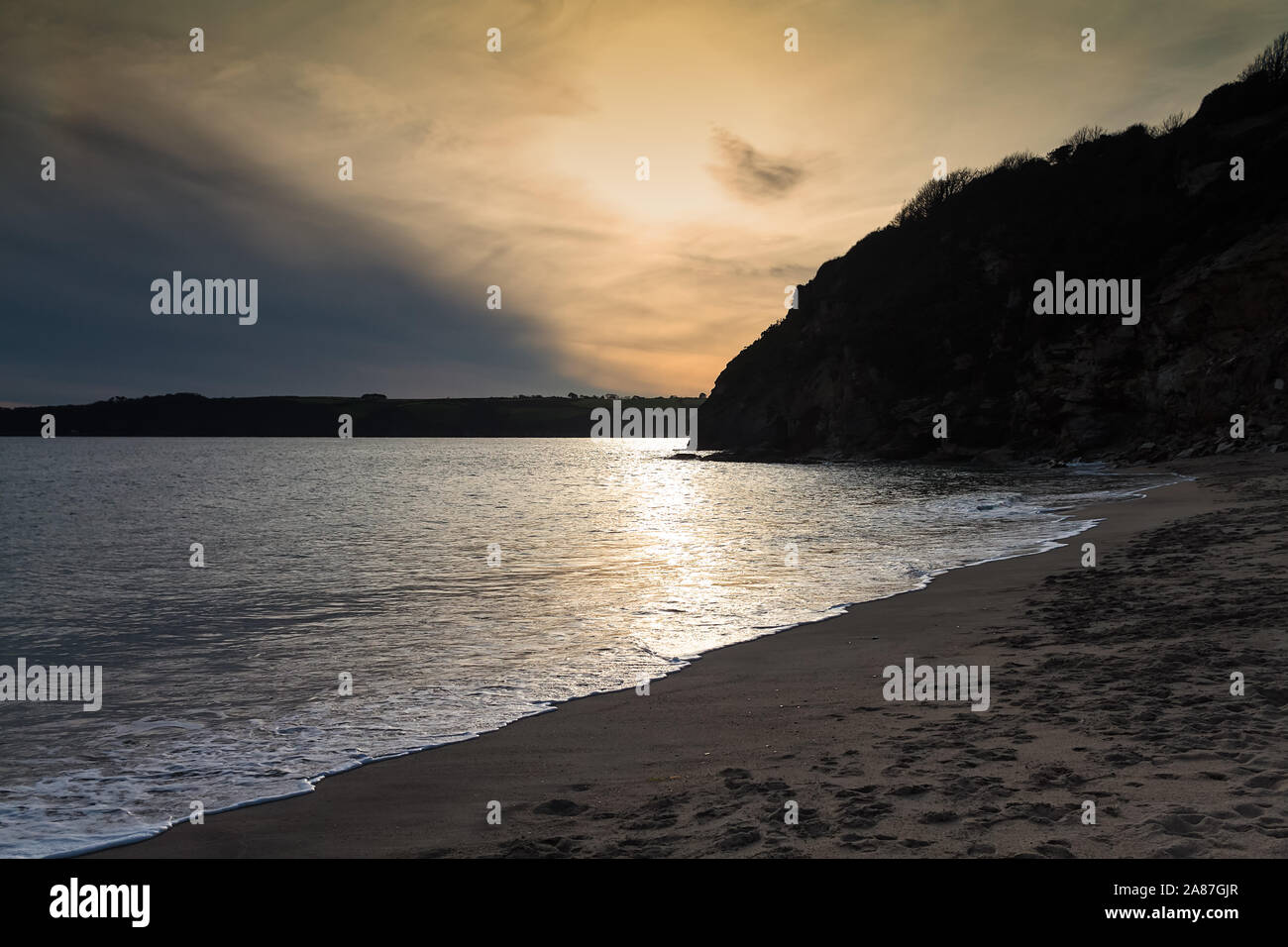 The sun sets behind the rocks at a beach in Cornwall, England. The ...