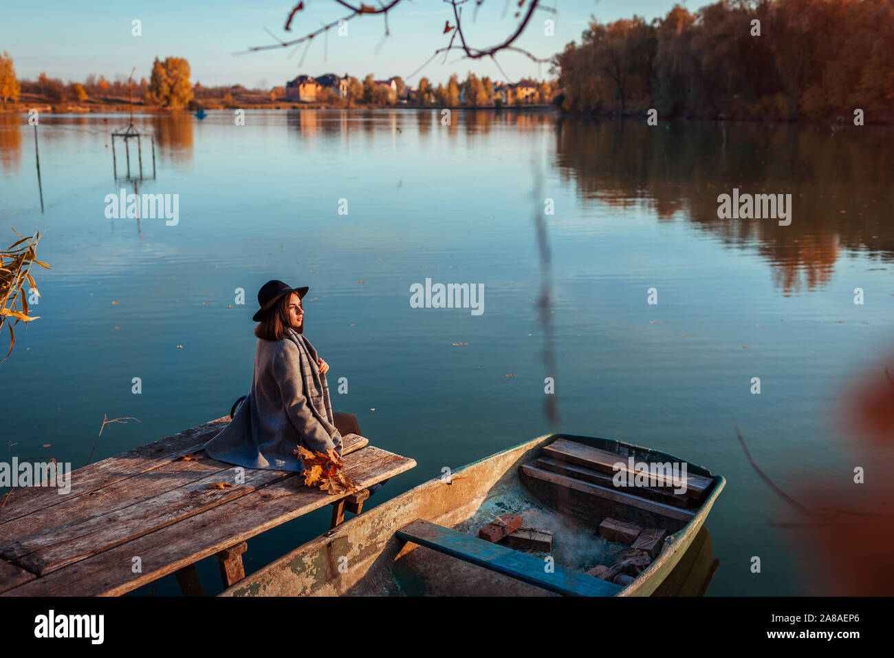 Woman sitting on lake pier by boat admiring autumn landscape and ...