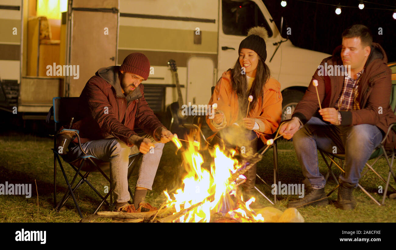 Friends relaxing around camp fire and roasting marshmallows on sticks ...