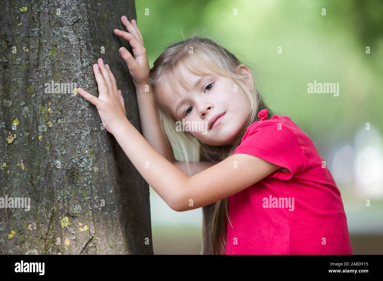 Portrait of a pretty little child girl standing near big tree trunk in ...