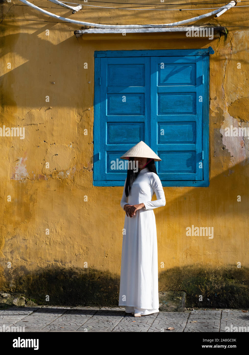 Young Vietnamese woman wearing a white ao dai and conical straw hat or non la. She is standing ...