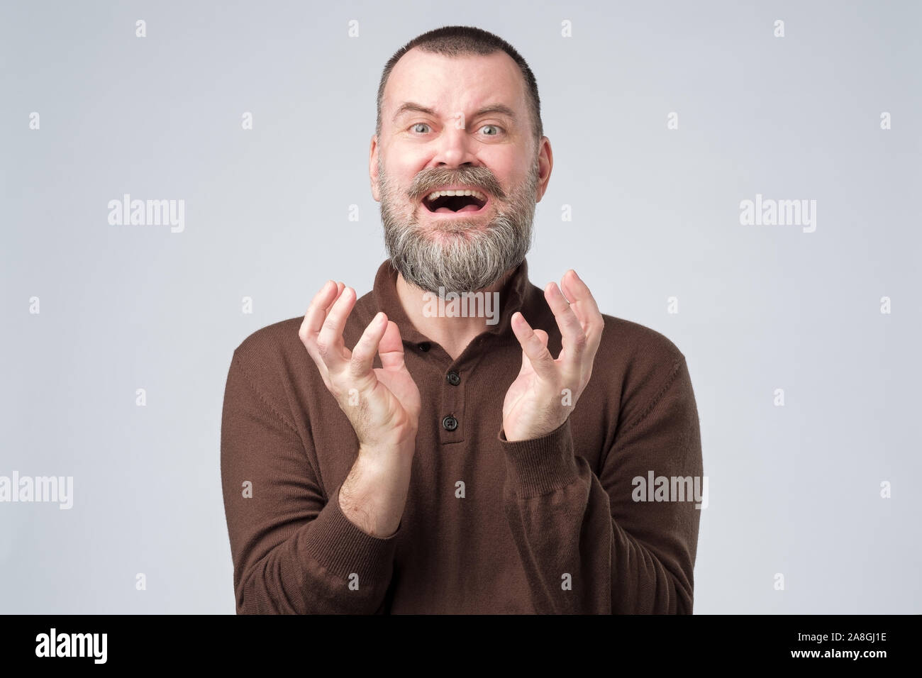 Mature scared bearded man in brown clothes squeezes fingers from horror ...