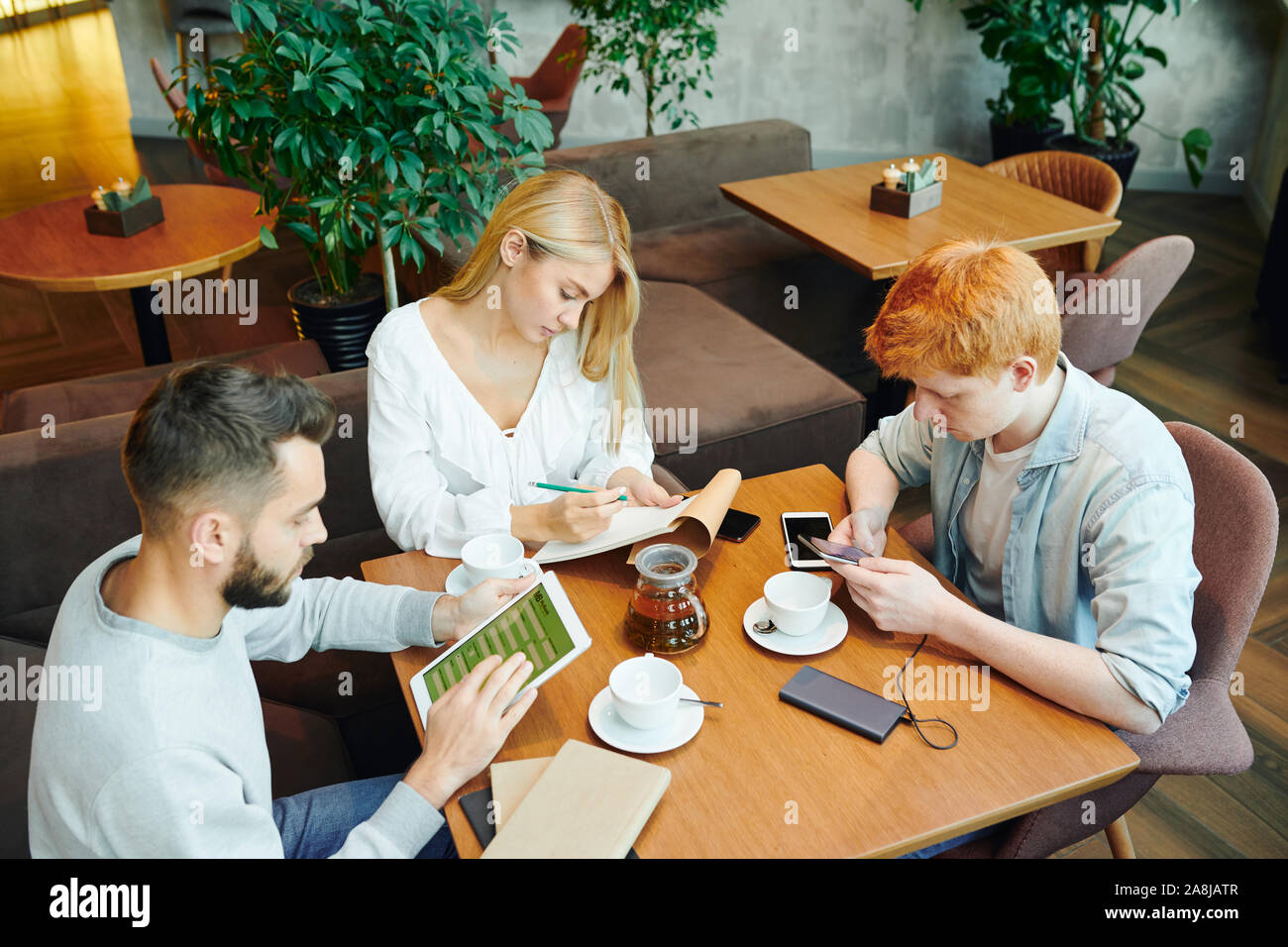 Two casual guys using gadgets while blonde girl making notes in notepad ...