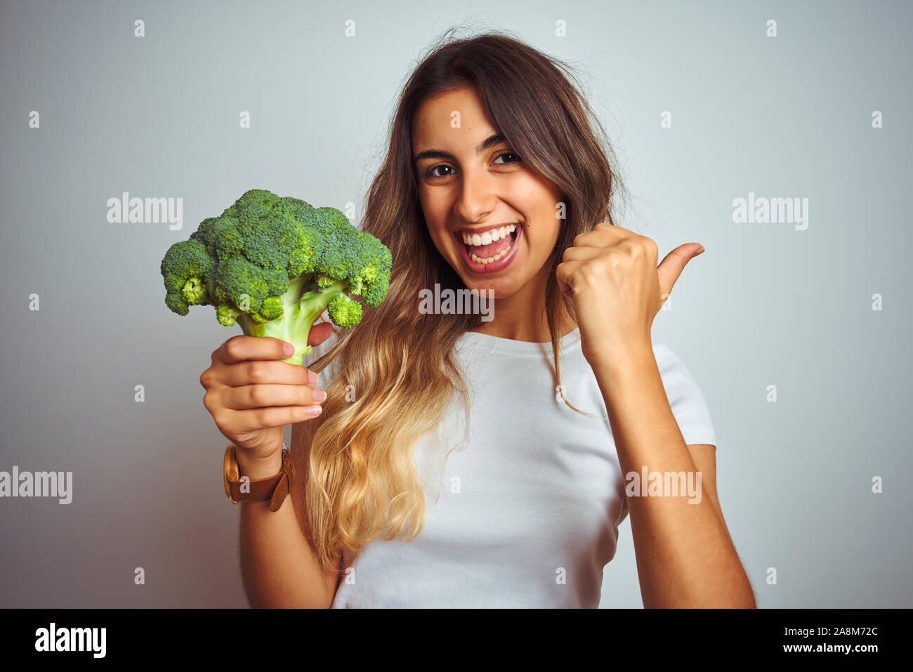 Young beautiful woman eating broccoli over grey isolated background ...
