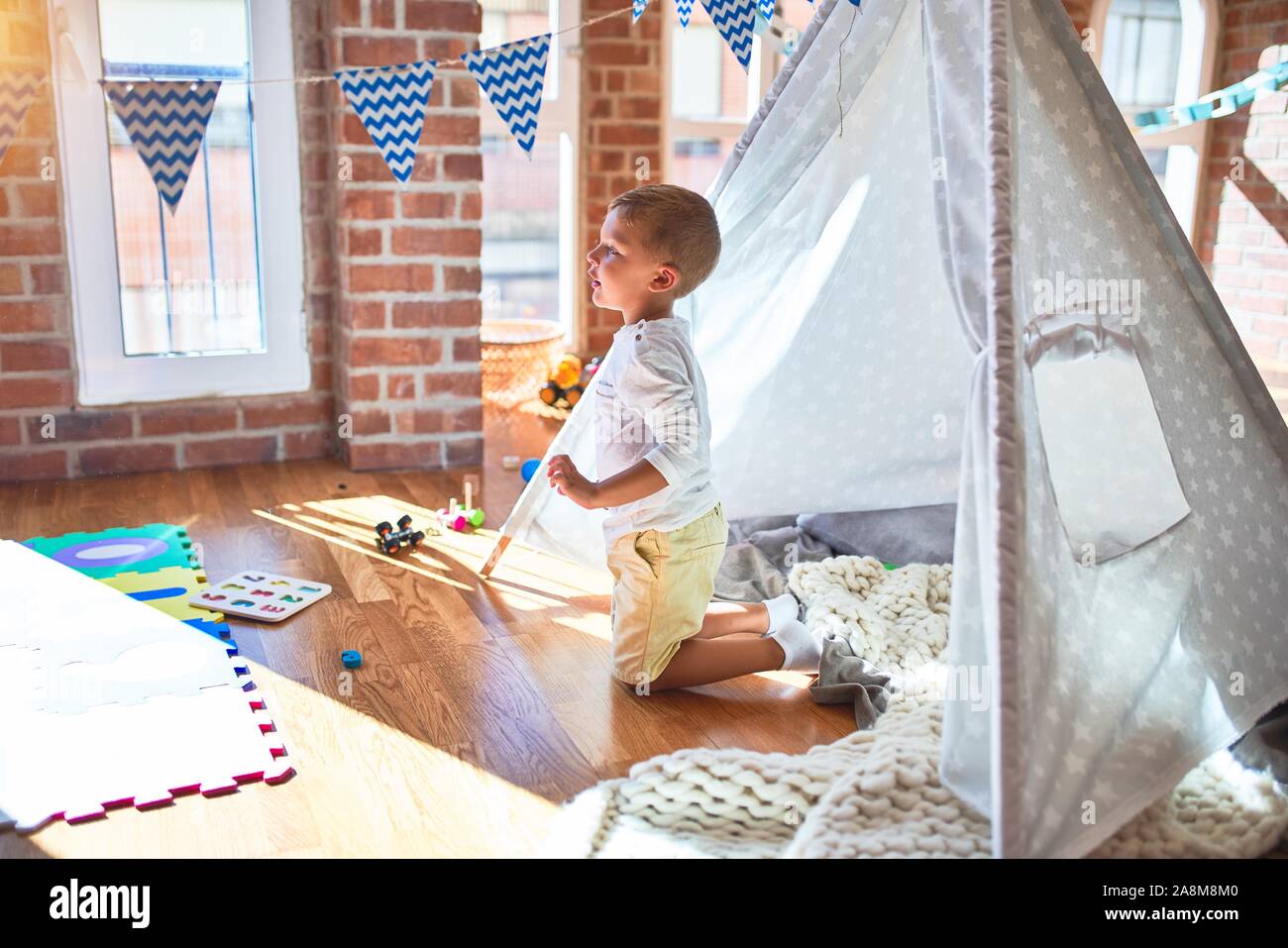 Beautiful blonde toddler sitting on the floor over blanket inside tipi ...