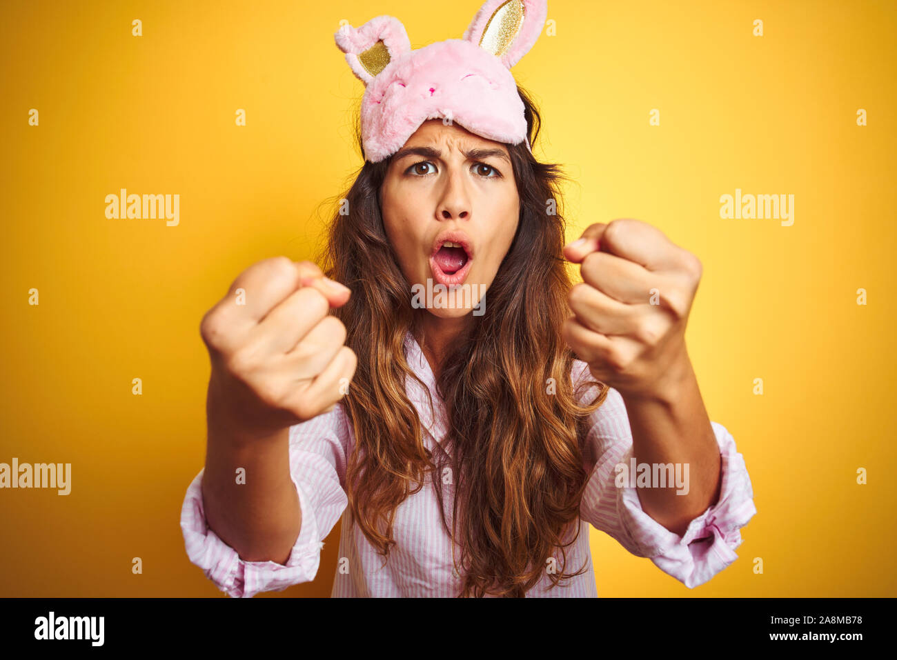 Young woman wearing pajama and sleep mask standing over yellow isolated ...