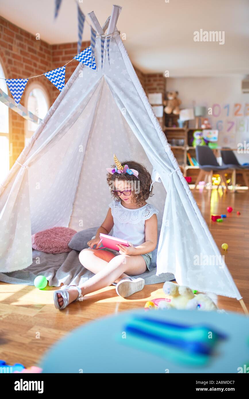 Beautiful toddler wearing glasses and unicorn diadem sitting on the floor inside tipi reading ...