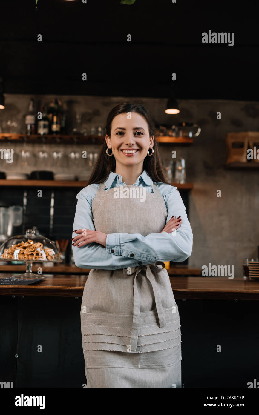 smiling barista in apron standing with crossed arms near bar counter ...