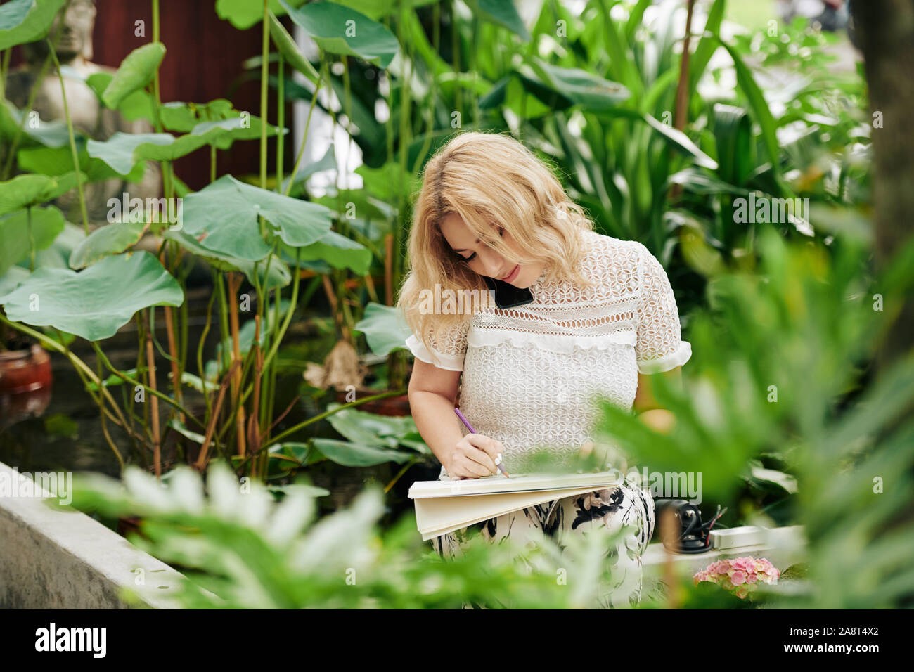 Creative young woman sitting in flower garden, talking on phone with ...