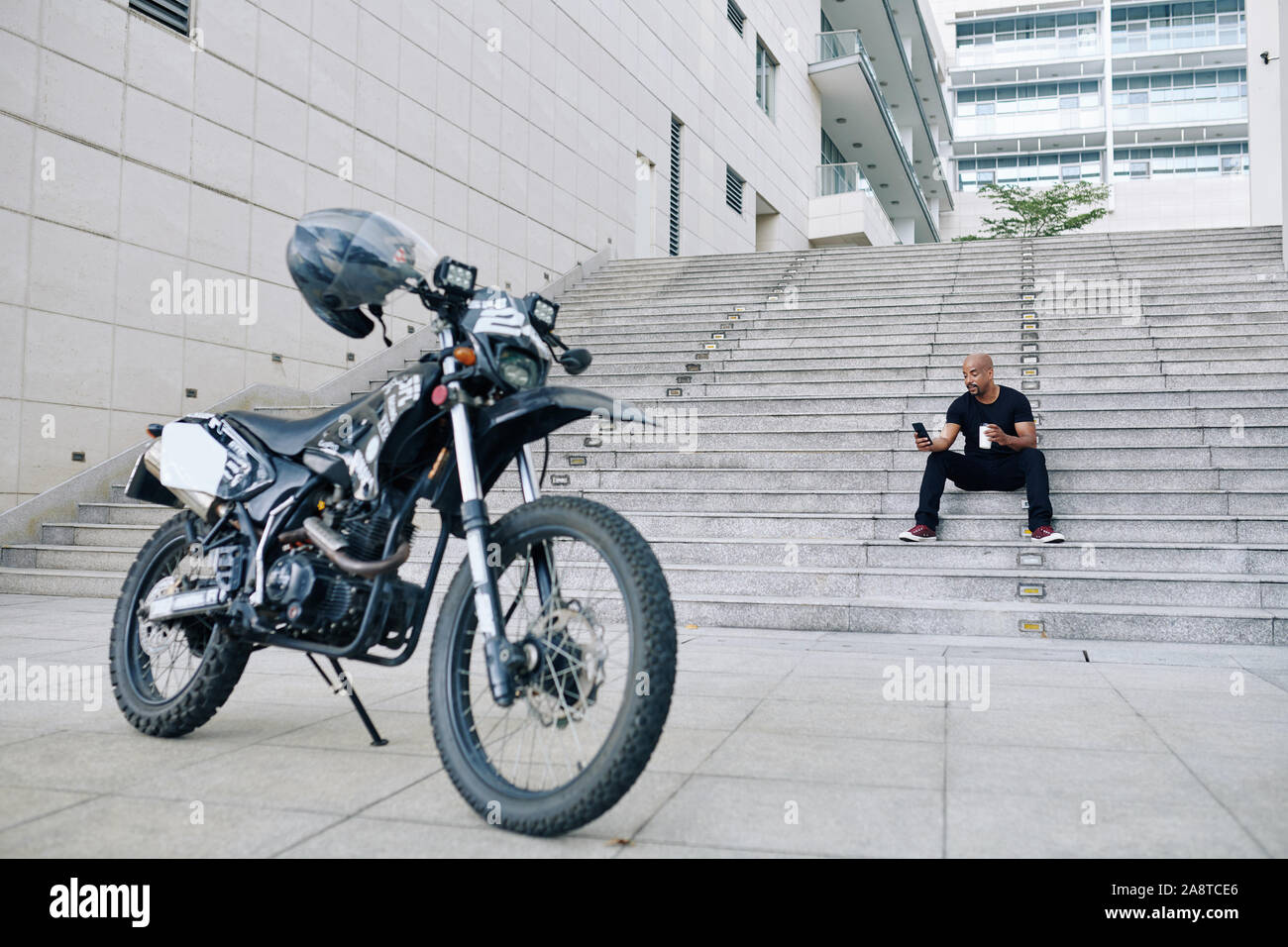 Handsome muscular motorcyclist sitting on steps, drinking take away ...