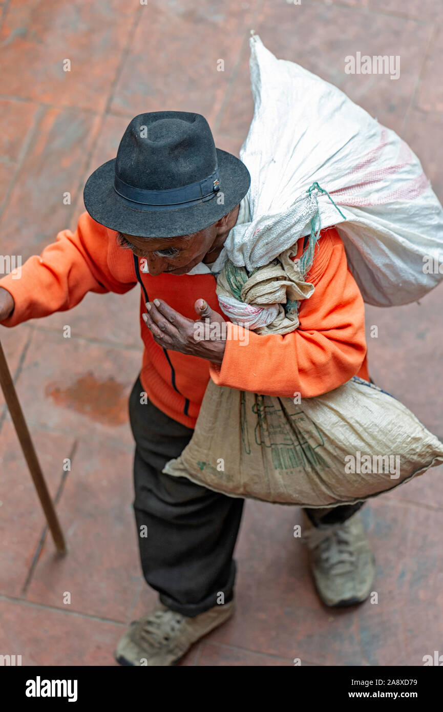 Elderly man wearing hat and orange jacket carries heavy sacks on his ...