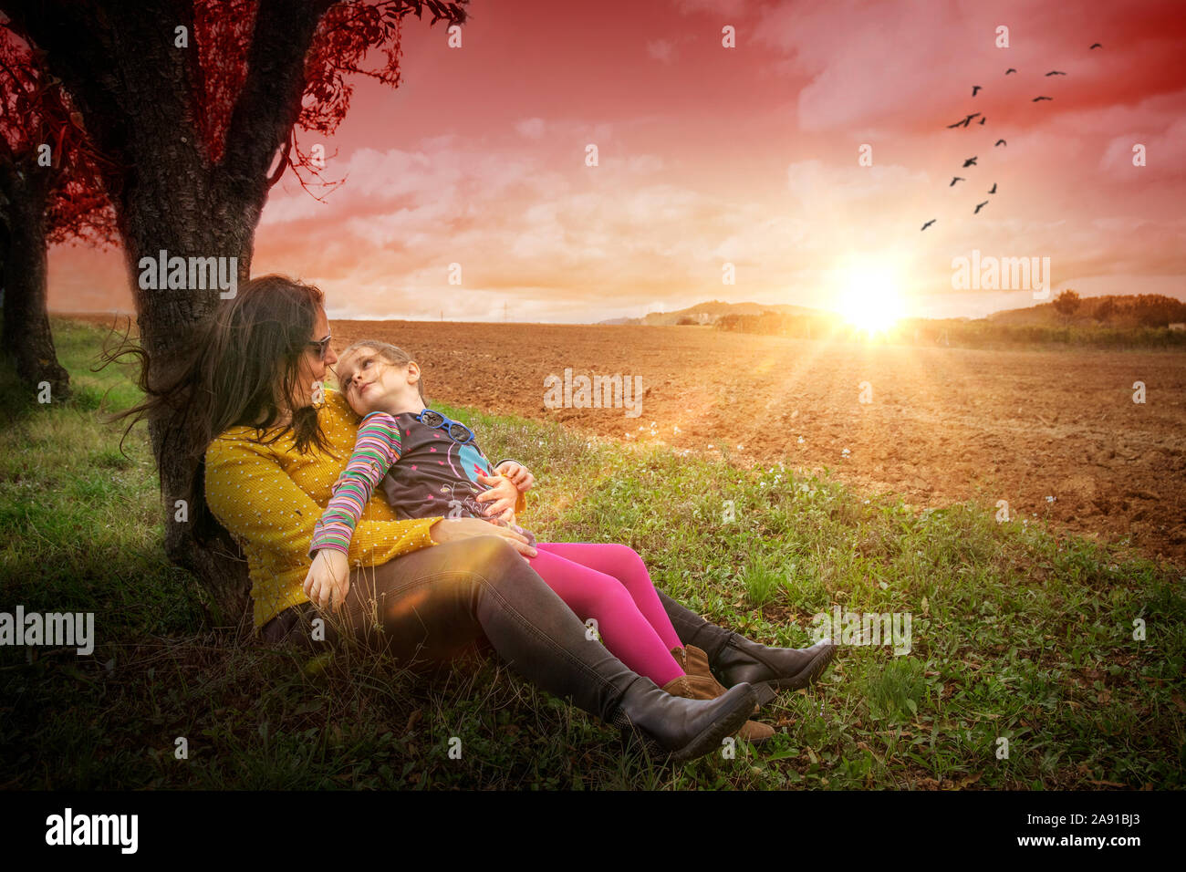 Mother and daughter sitting under a tree in the meadow at sunset. Empty ...