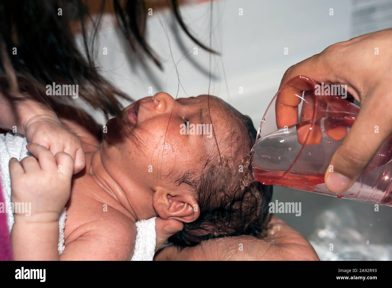 Glass with water being poured over head of crying two week old baby to ...