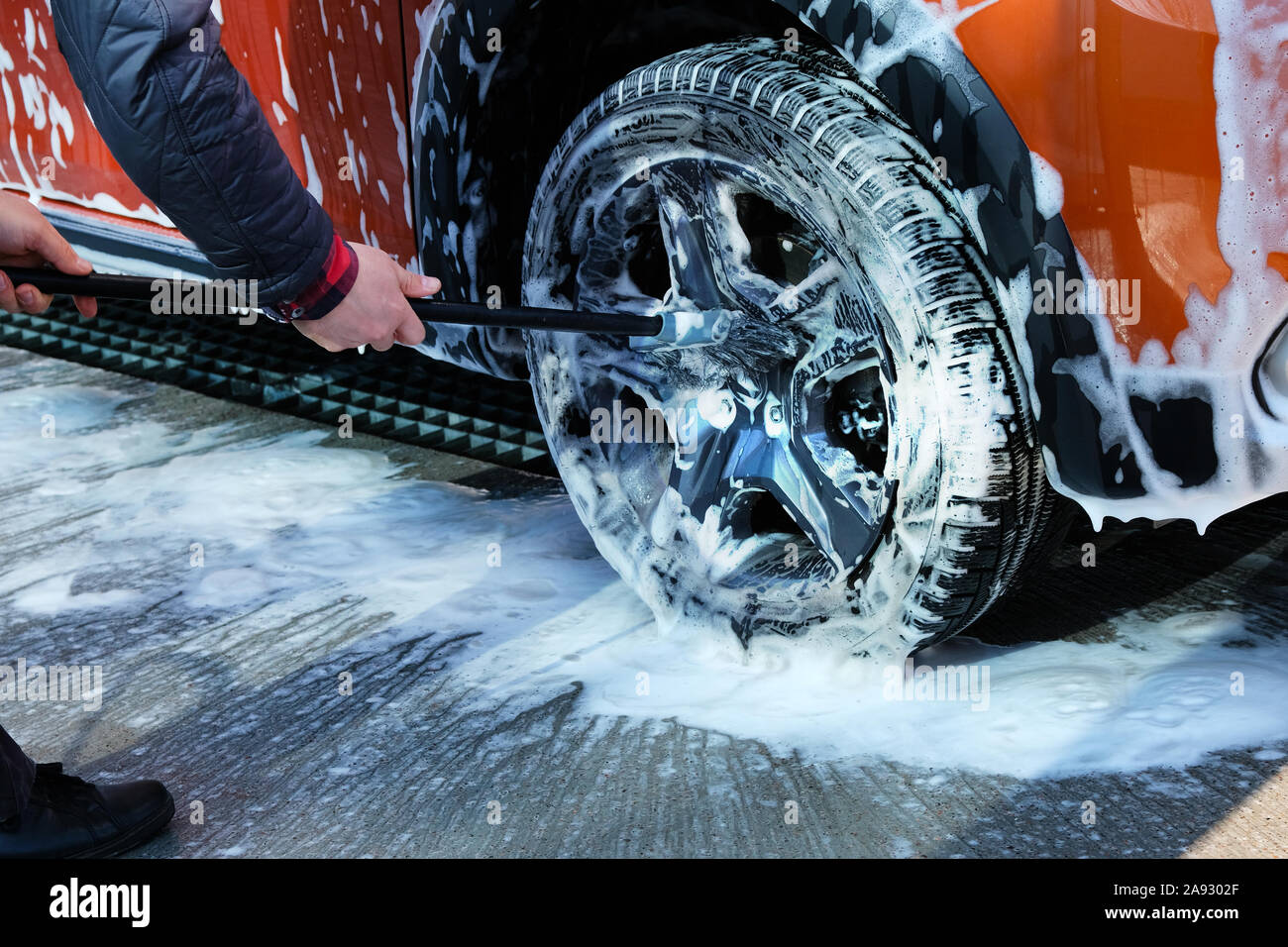 Man washes black wheel of his orange car with brush. Cleaning with soap ...