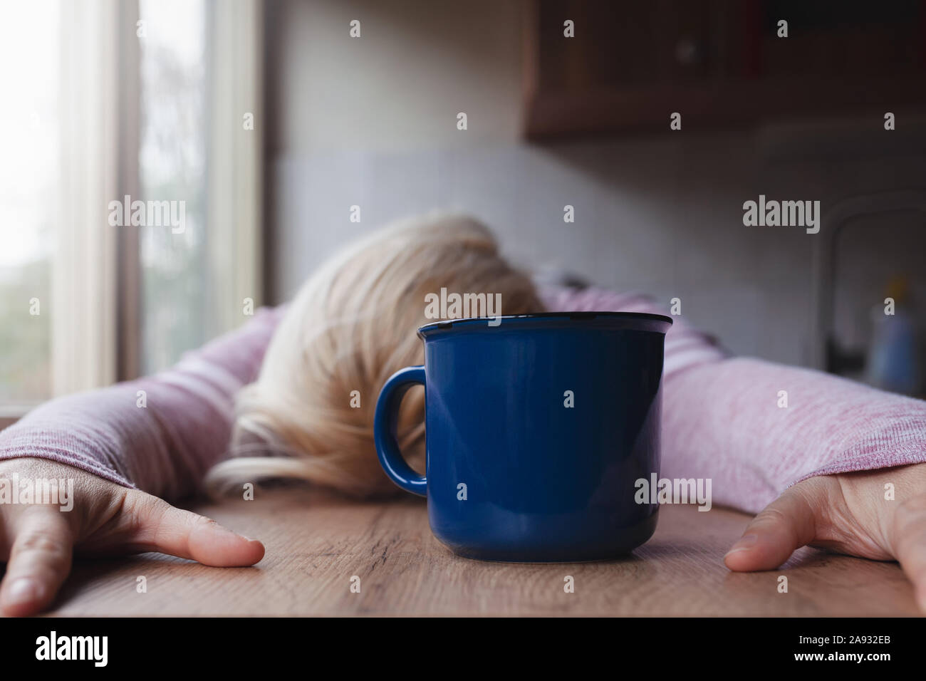Blonde woman resting her head on a kitchen table, blue cup of coffee in ...