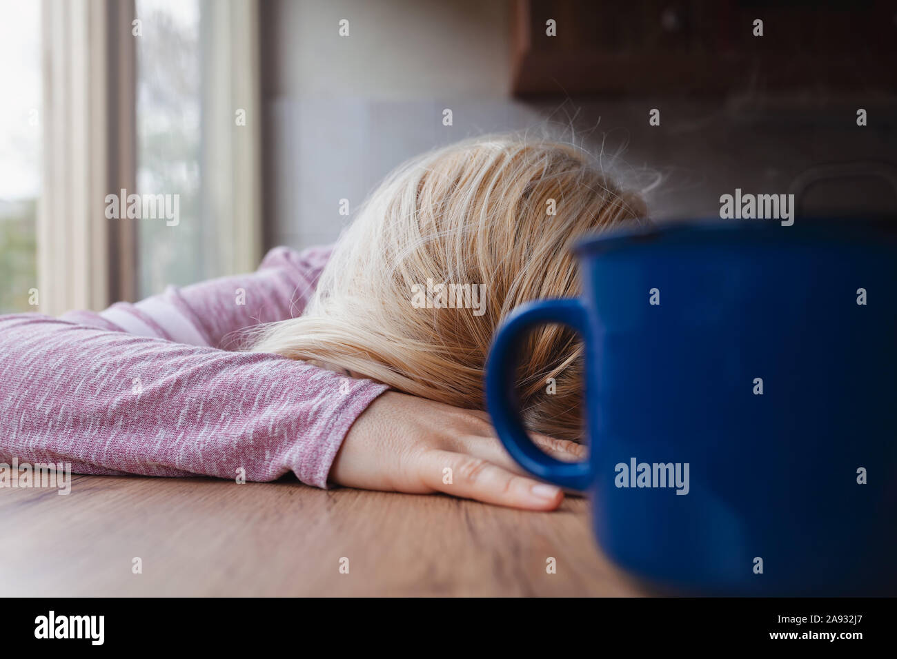 Blonde woman resting her head on a kitchen table, blue cup of coffee in ...