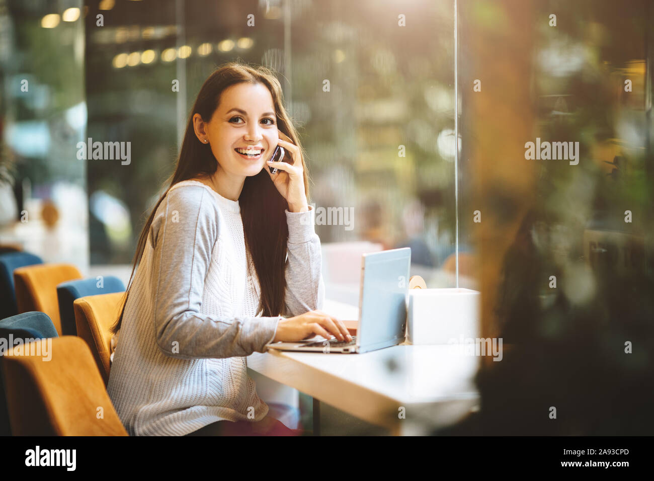 Beautiful Caucasian woman uses Phone while sitting with portable net-book in modern cafe bar ...