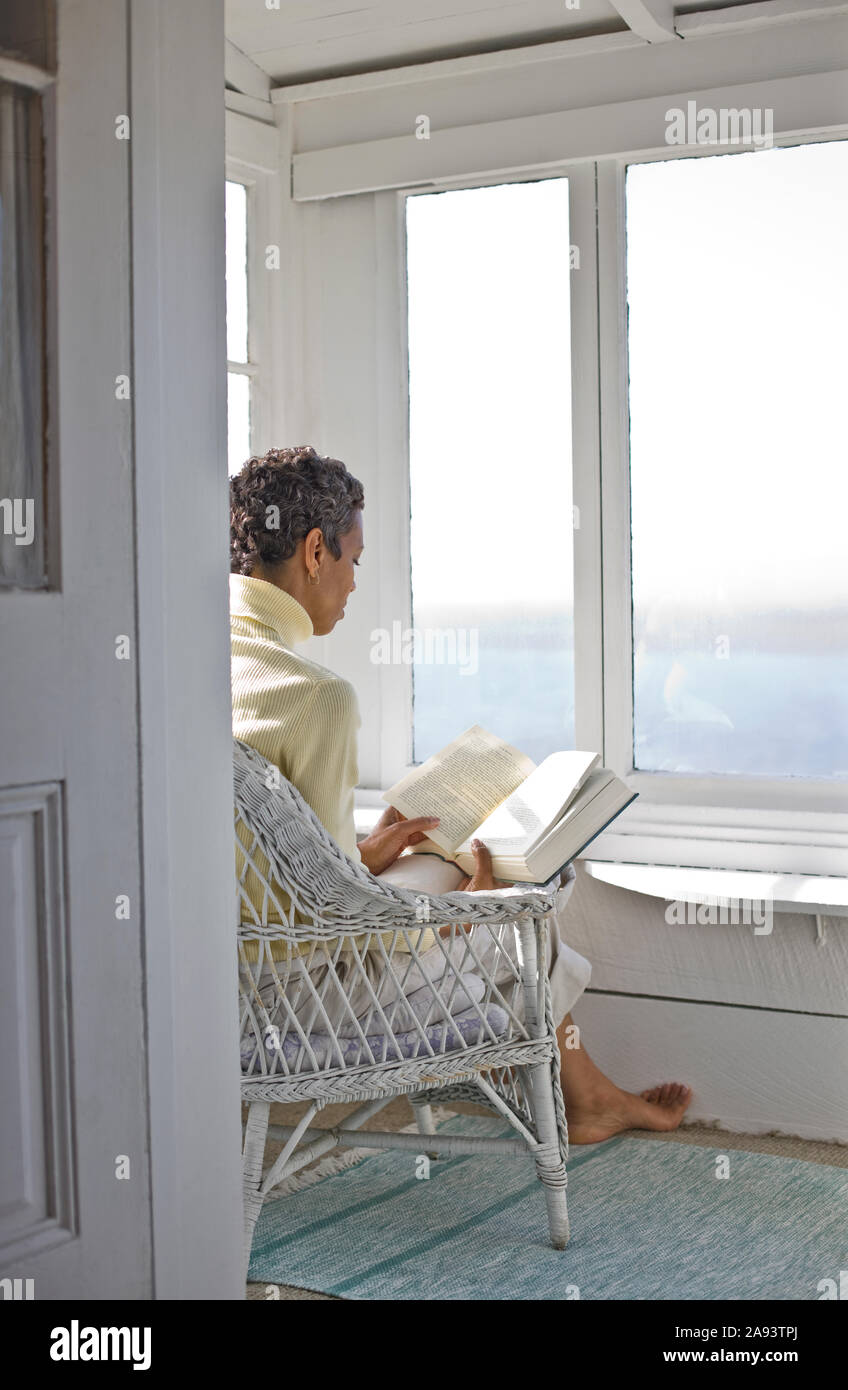 Mid-adult woman sitting in a wicker chair reading a book in a sunny ...