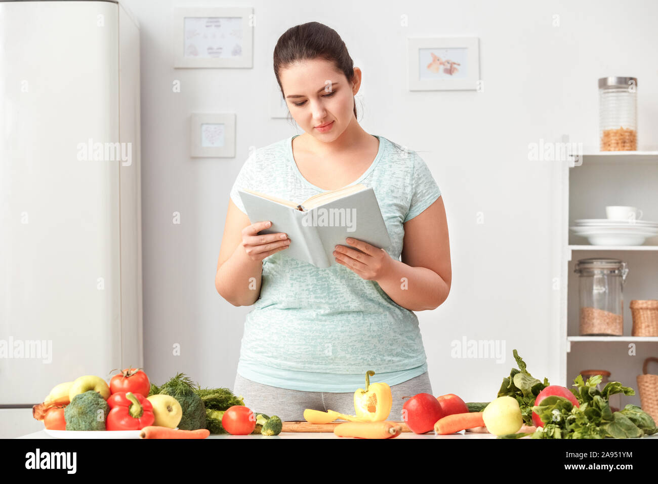Cooking Meal. Chubby girl standing in kitchen reading book of recipes ...