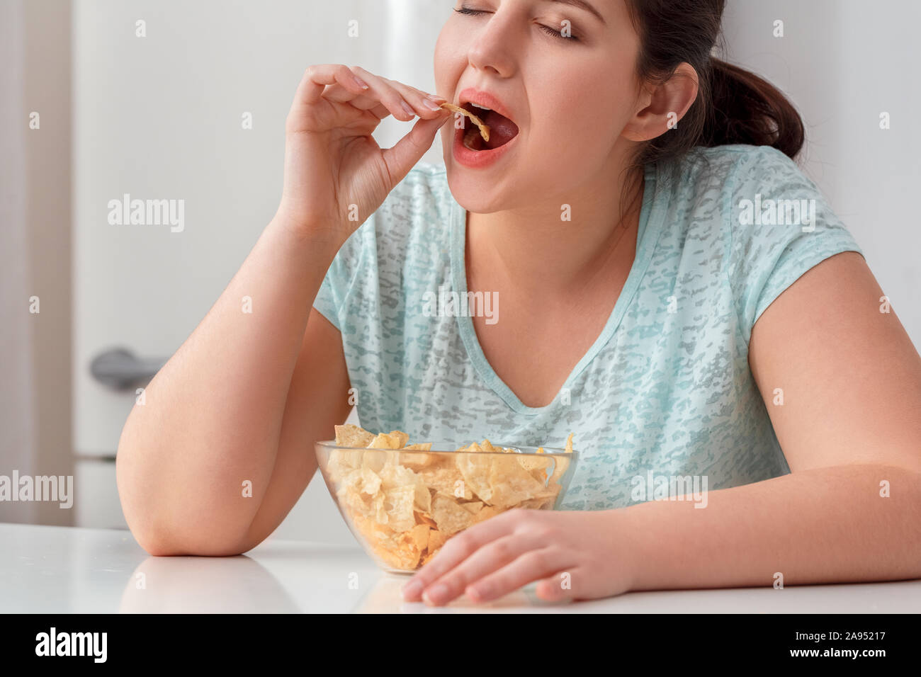 Breaking Diet. Chubby girl sitting at kitchen table eating chips closed ...