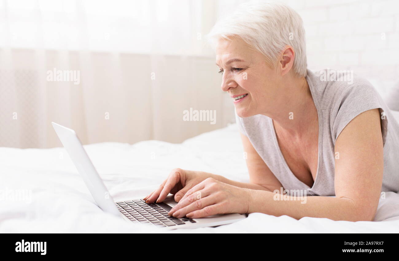Senior lady browsing on laptop, resting on bed, empty space Stock Photo ...