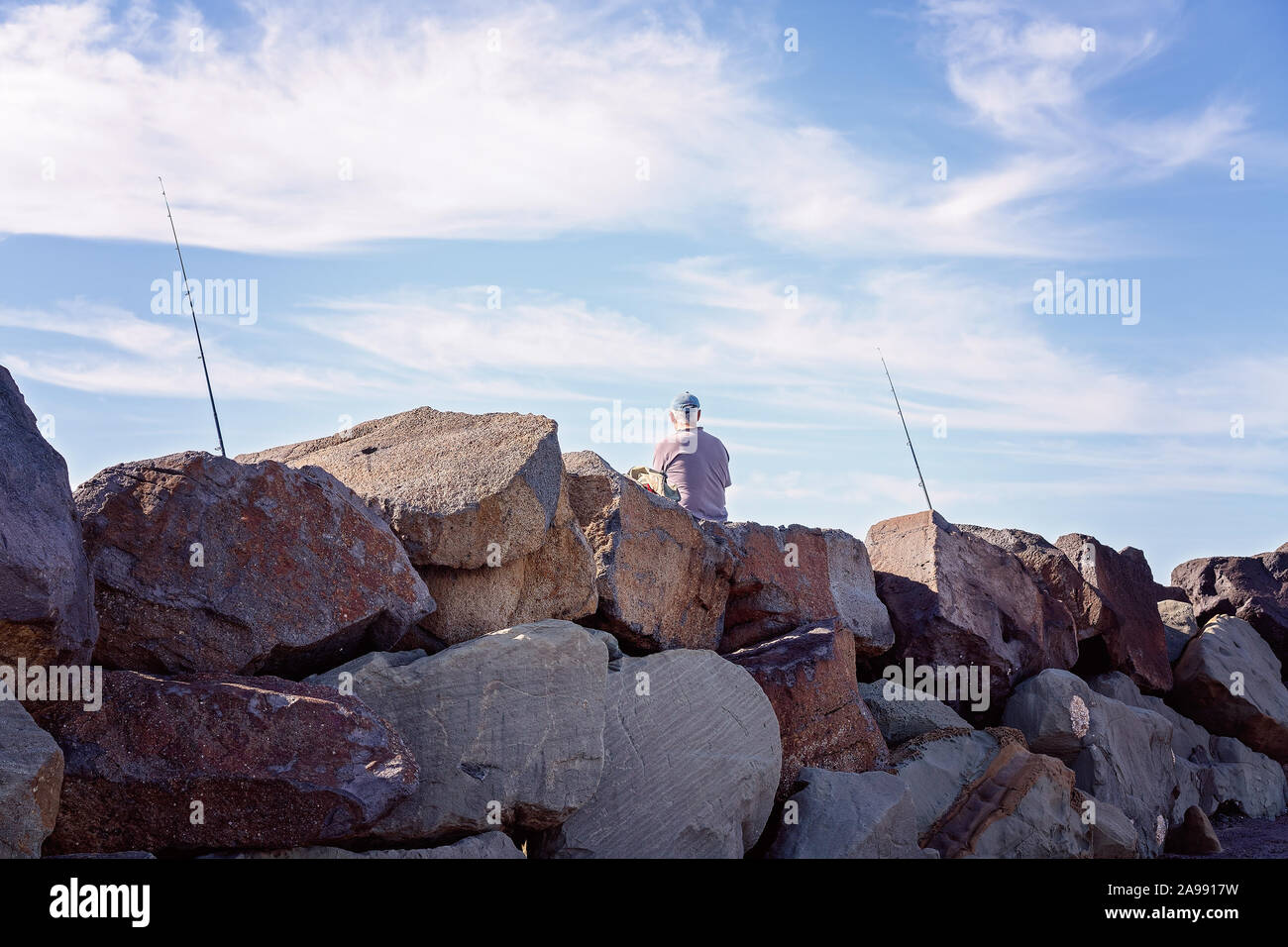 Unidentified man fishing with two rods out sitting on breakwater rocks ...