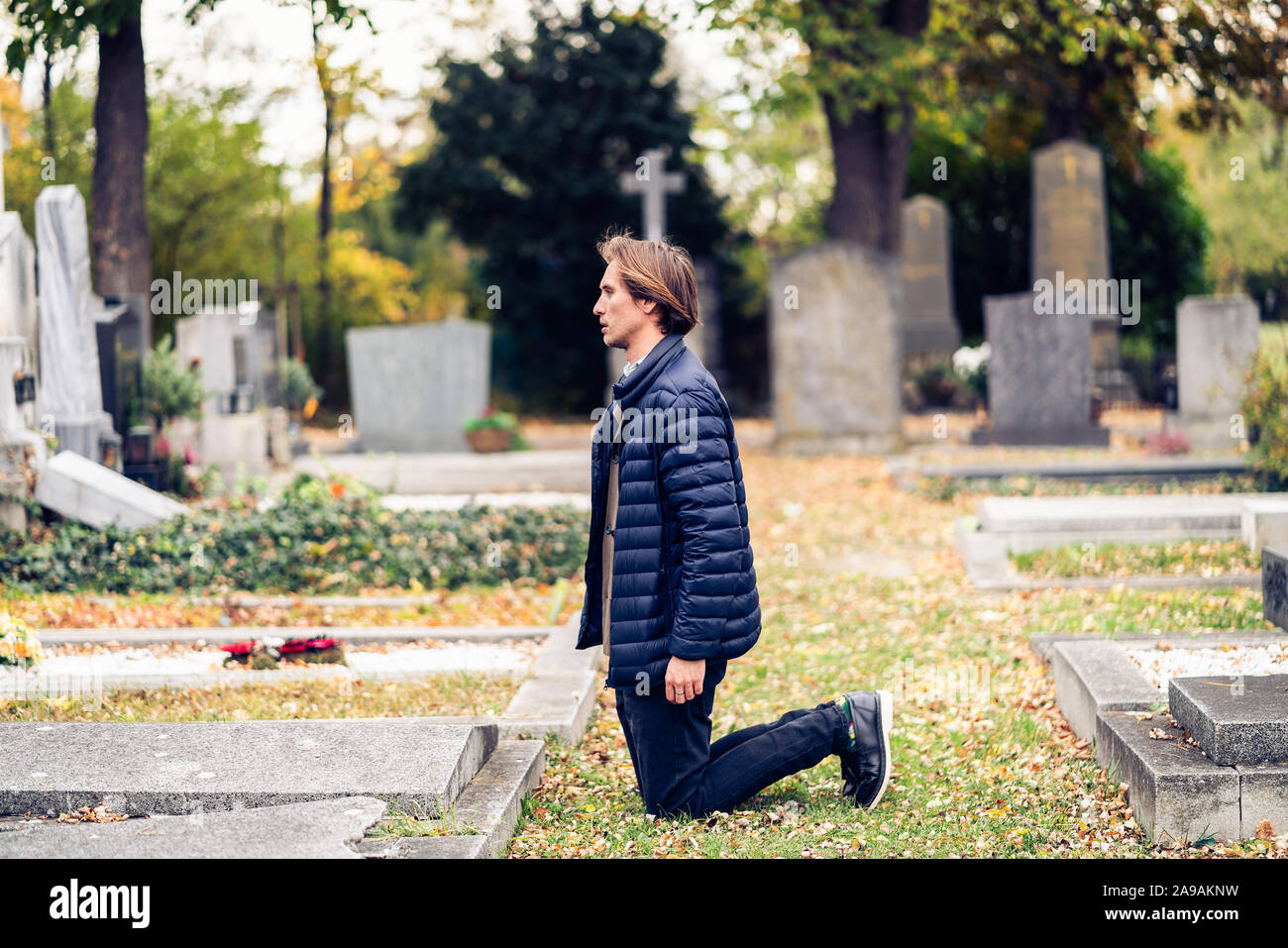Mourning young man kneeling in front of a grave on a cemetery during a ...