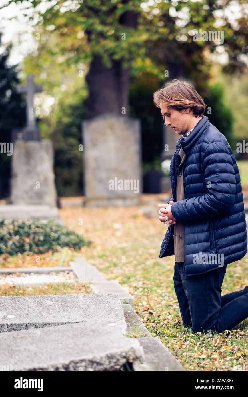 Mourning young man kneeling in front of a grave on a cemetery during a ...