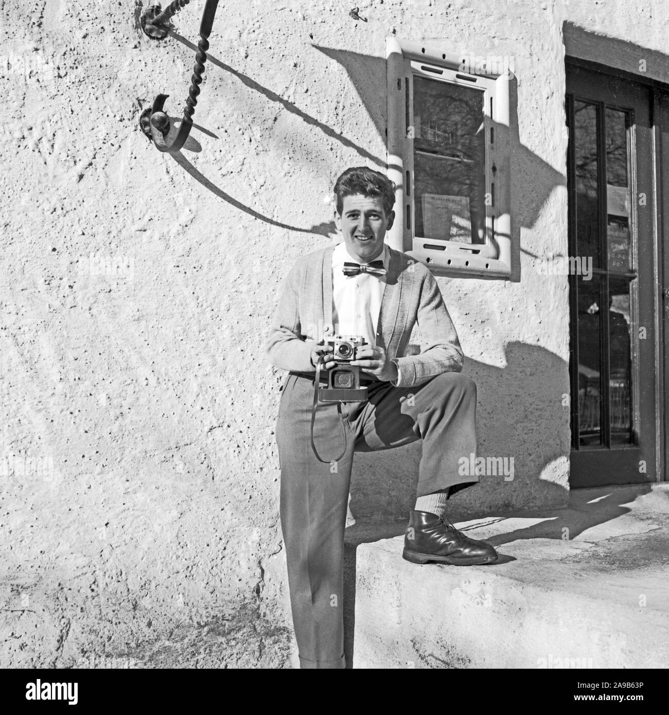 A young man posing with a camera in front of a house, Germany 1957 ...
