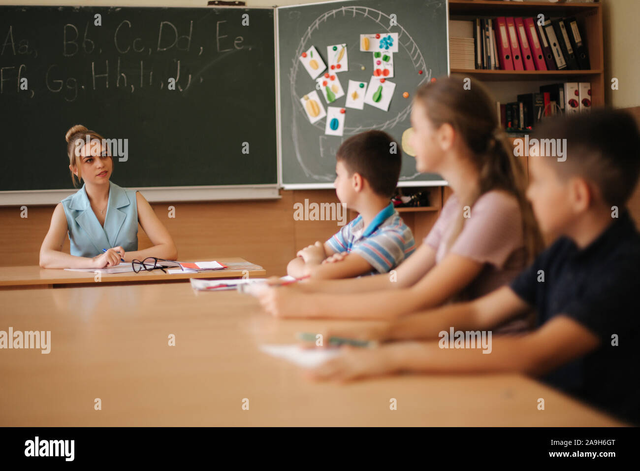Pretty teacher in classroom sitting at the desk and asking children ...