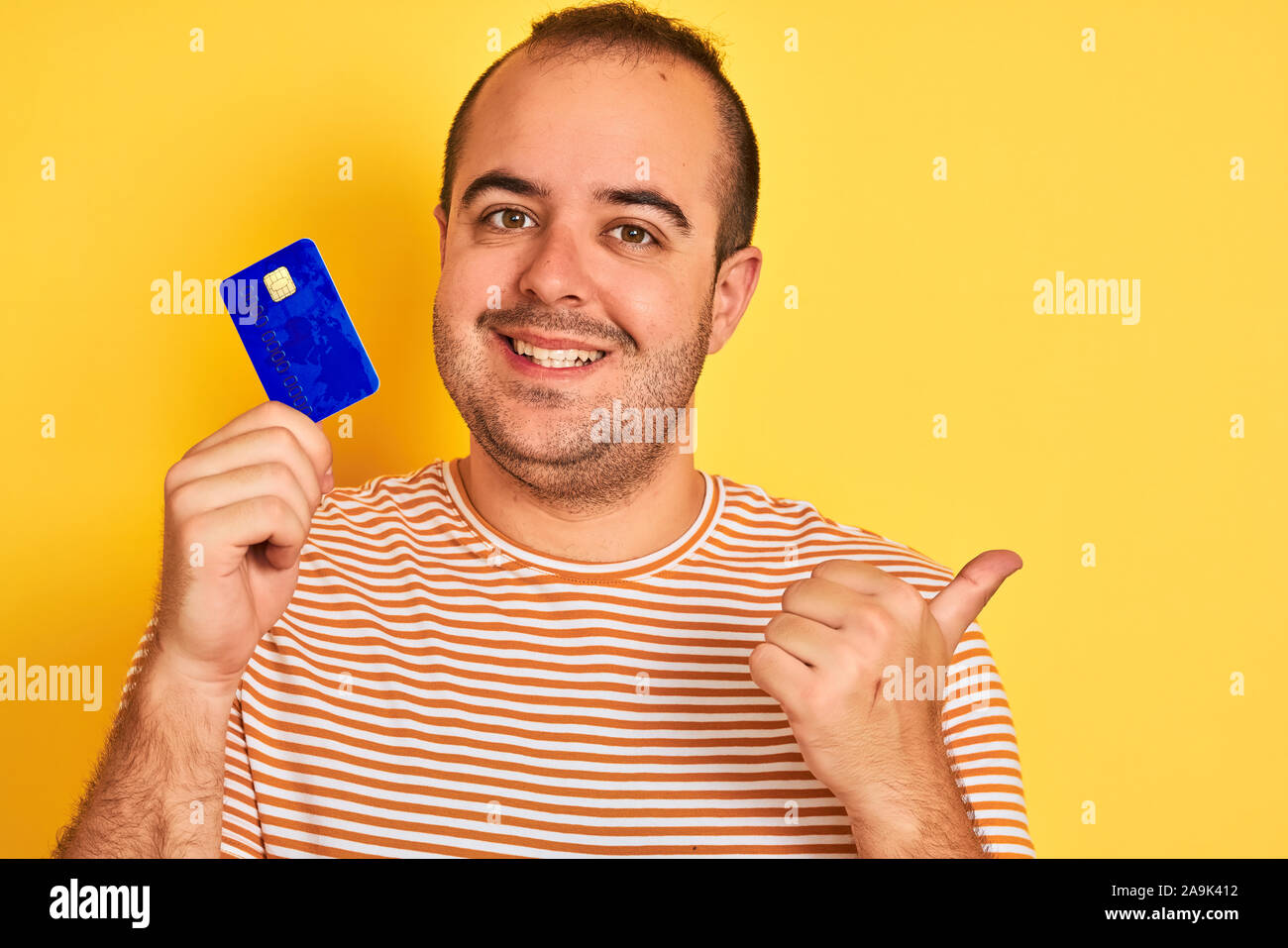 Young man holding blue credit card standing over isolated yellow ...