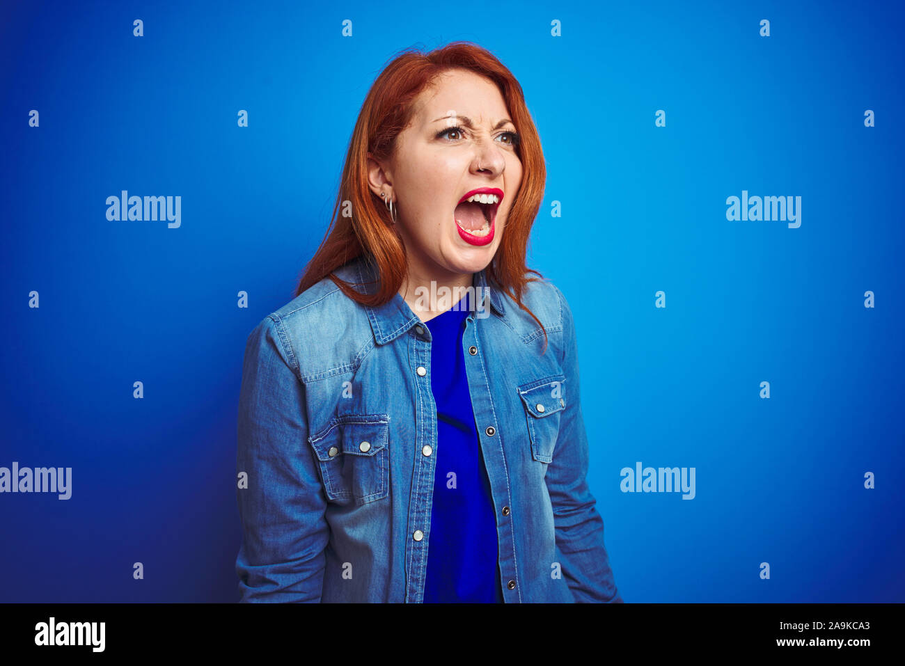 Young beautiful redhead woman wearing denim shirt standing over blue ...