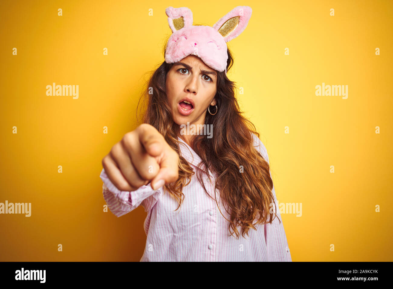 Young woman wearing pajama and sleep mask standing over yellow isolated ...
