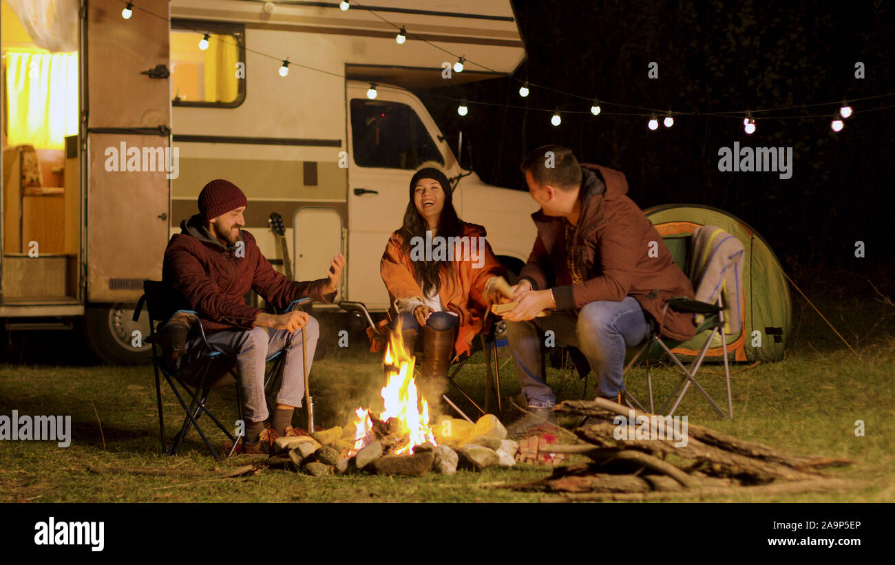 Group of close friends laughing together around camp fire. Retro camper ...