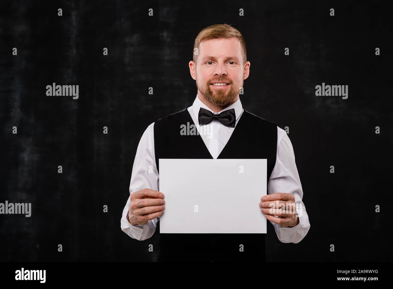 Happy elegant waiter in black waistcoat and bowtie holding blank paper ...