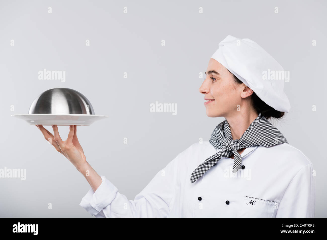 Pretty young smiling chef in white uniform holding cloche in front of ...