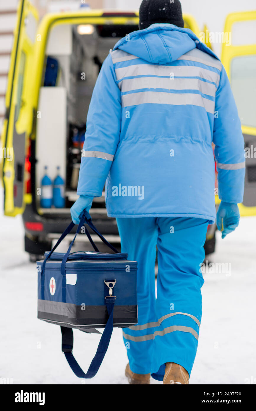 Rear view of paramedic in blue workwear and gloves carrying first aid ...