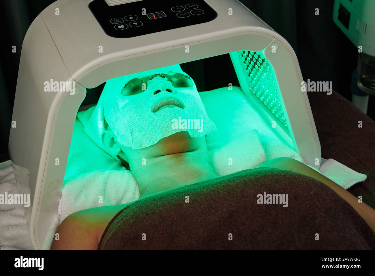 Young woman with sheet mask on her face having LED light treatment in ...