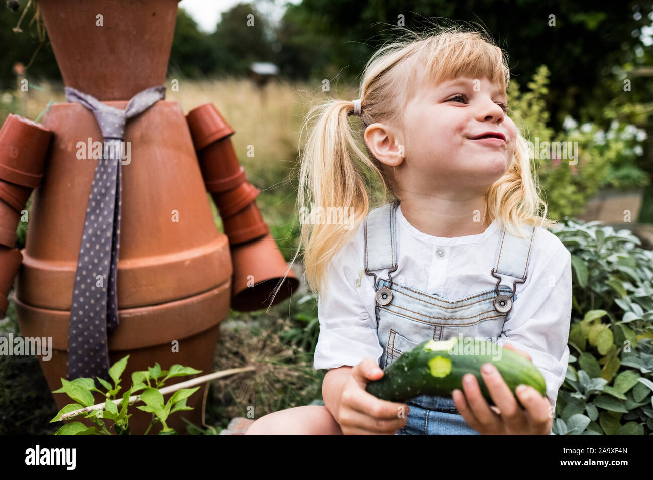 Blond girl sitting in garden next to terracotta scarecrow, eating ...