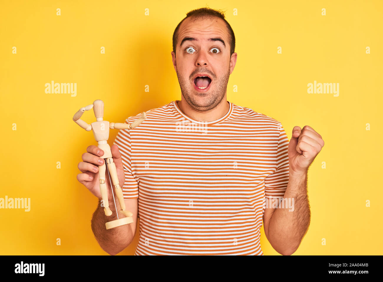 Young man holding figure of art dummy standing over isolated yellow ...