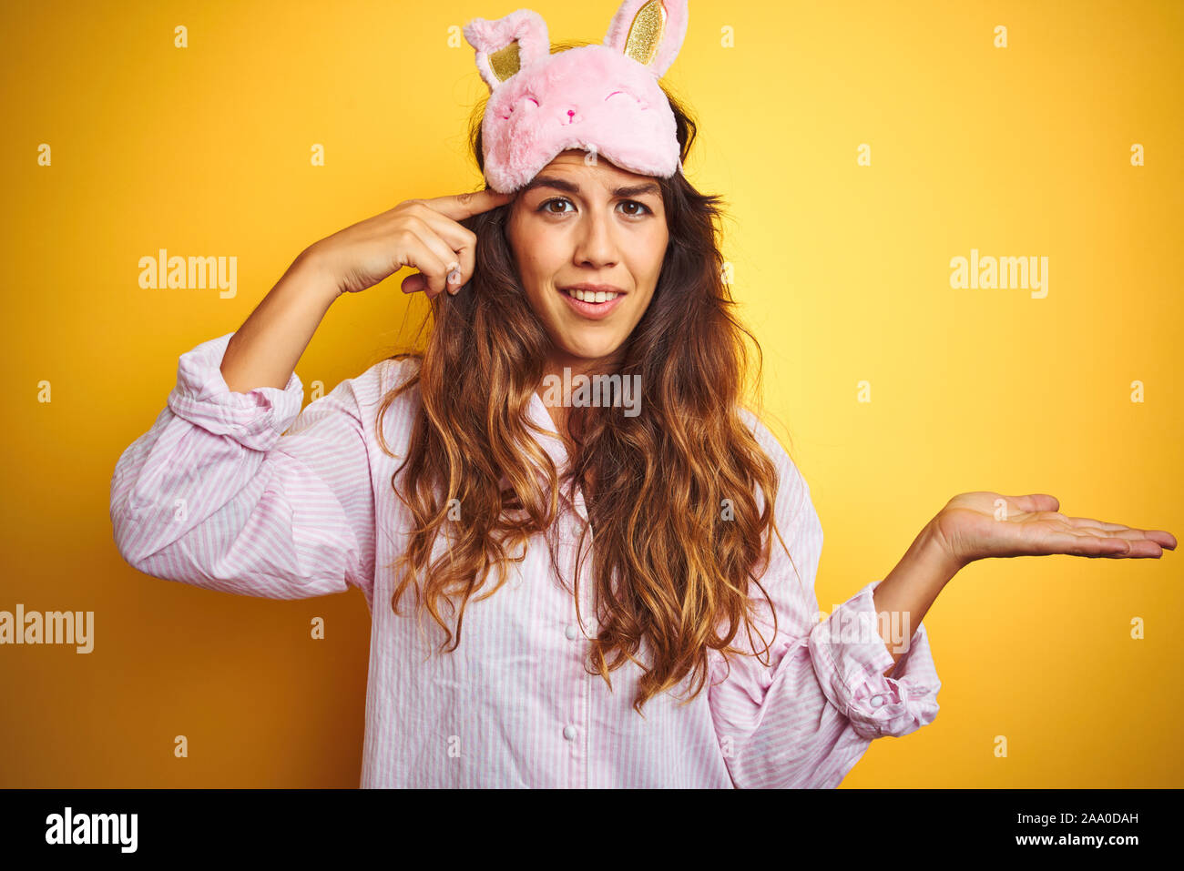 Young woman wearing pajama and sleep mask standing over yellow isolated ...