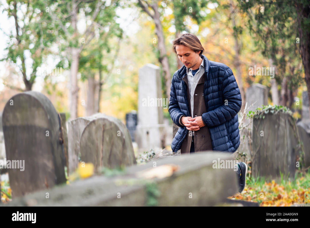 Mourning young man kneeling in front of a grave on a cemetery during a ...