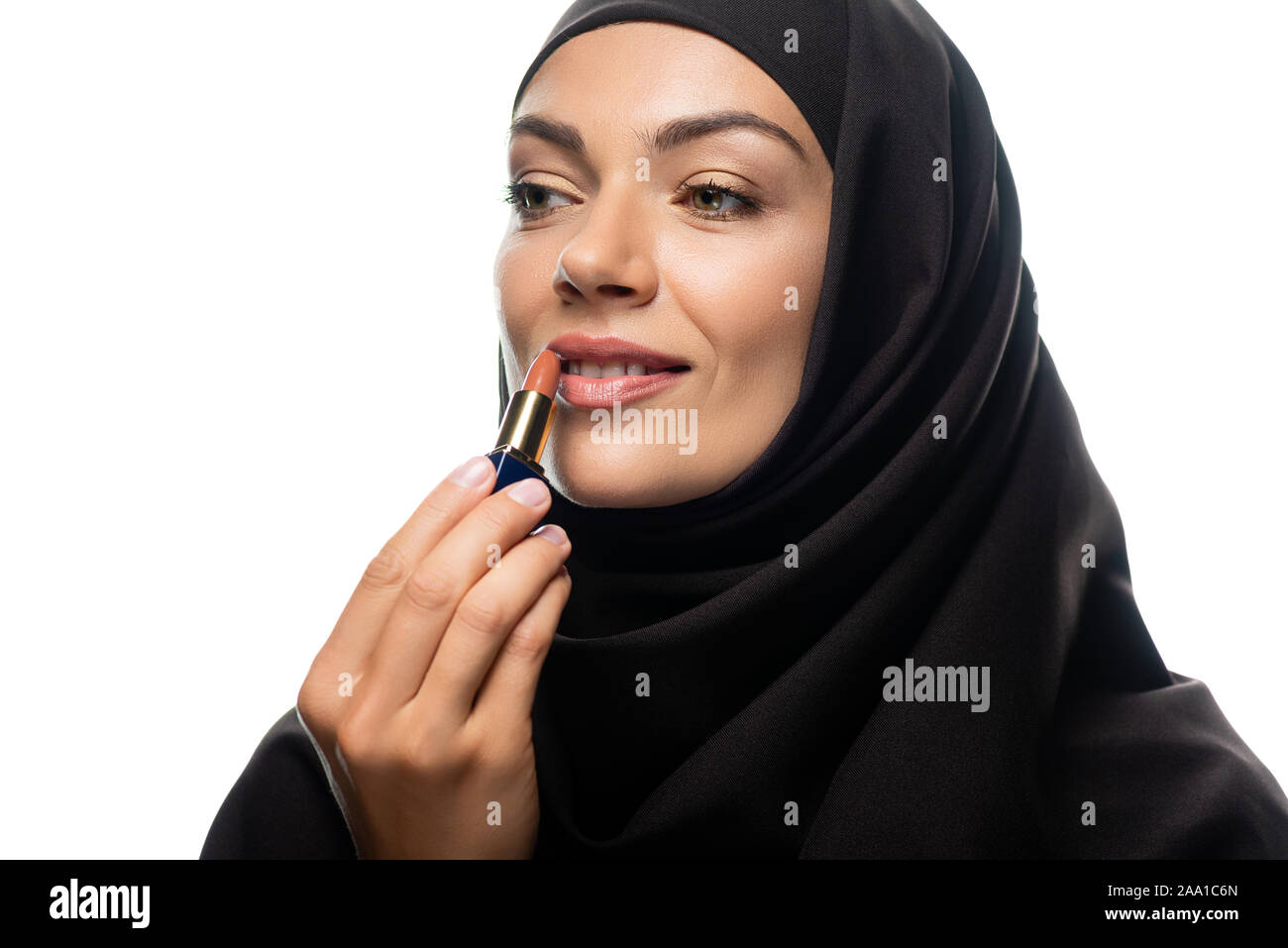 smiling young Muslim woman in hijab applying beige lipstick isolated on ...