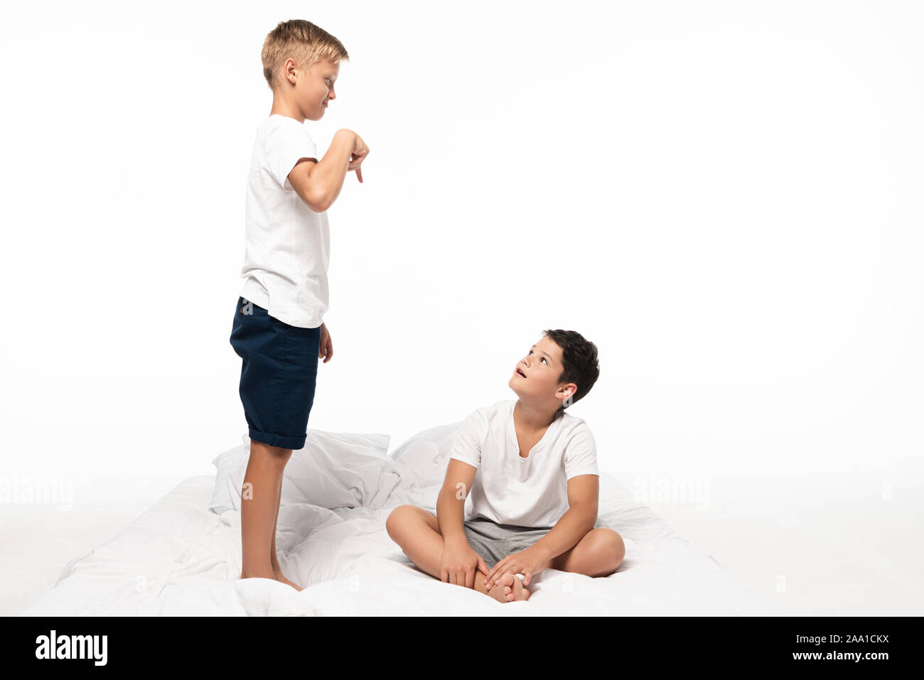 boy pointing with finger at brother sitting on bed isolated on white ...