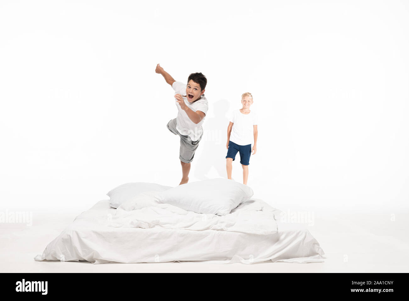 excited boy levitating over bed while smiling brother standing on white ...