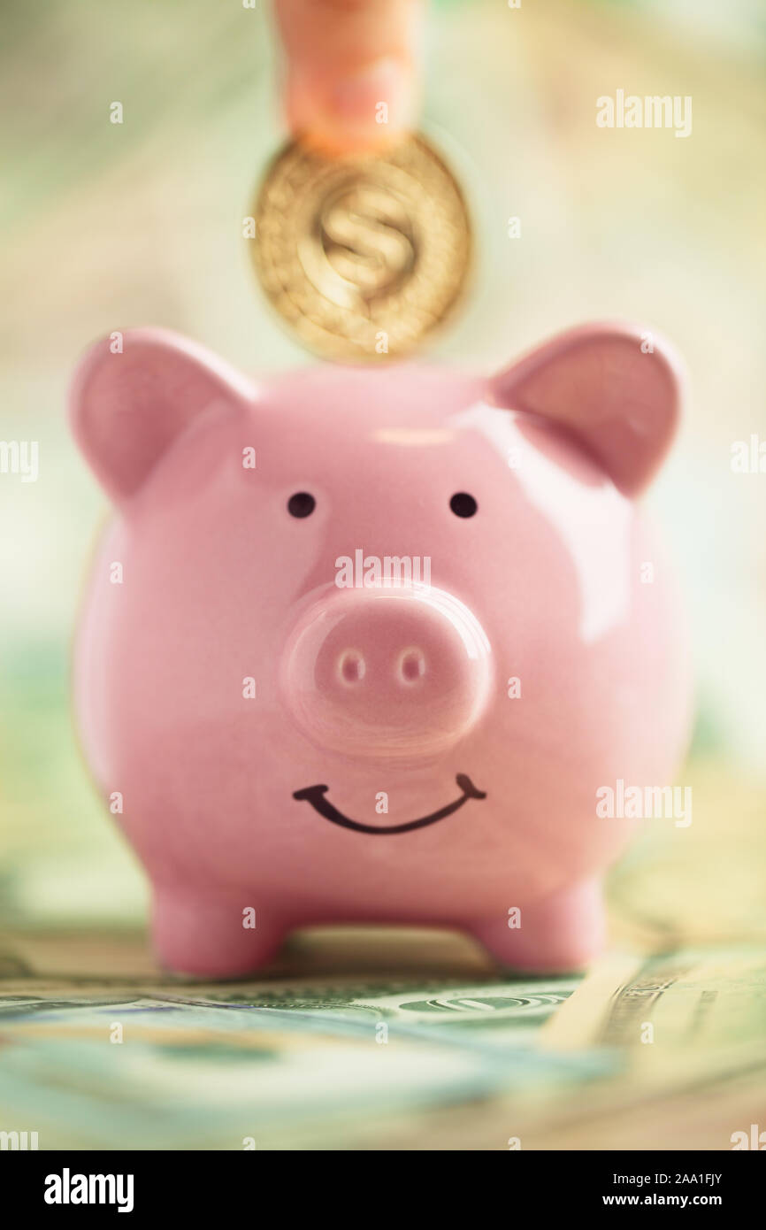 Depositing gold coin in smiling pink Piggybank on piles of money ...